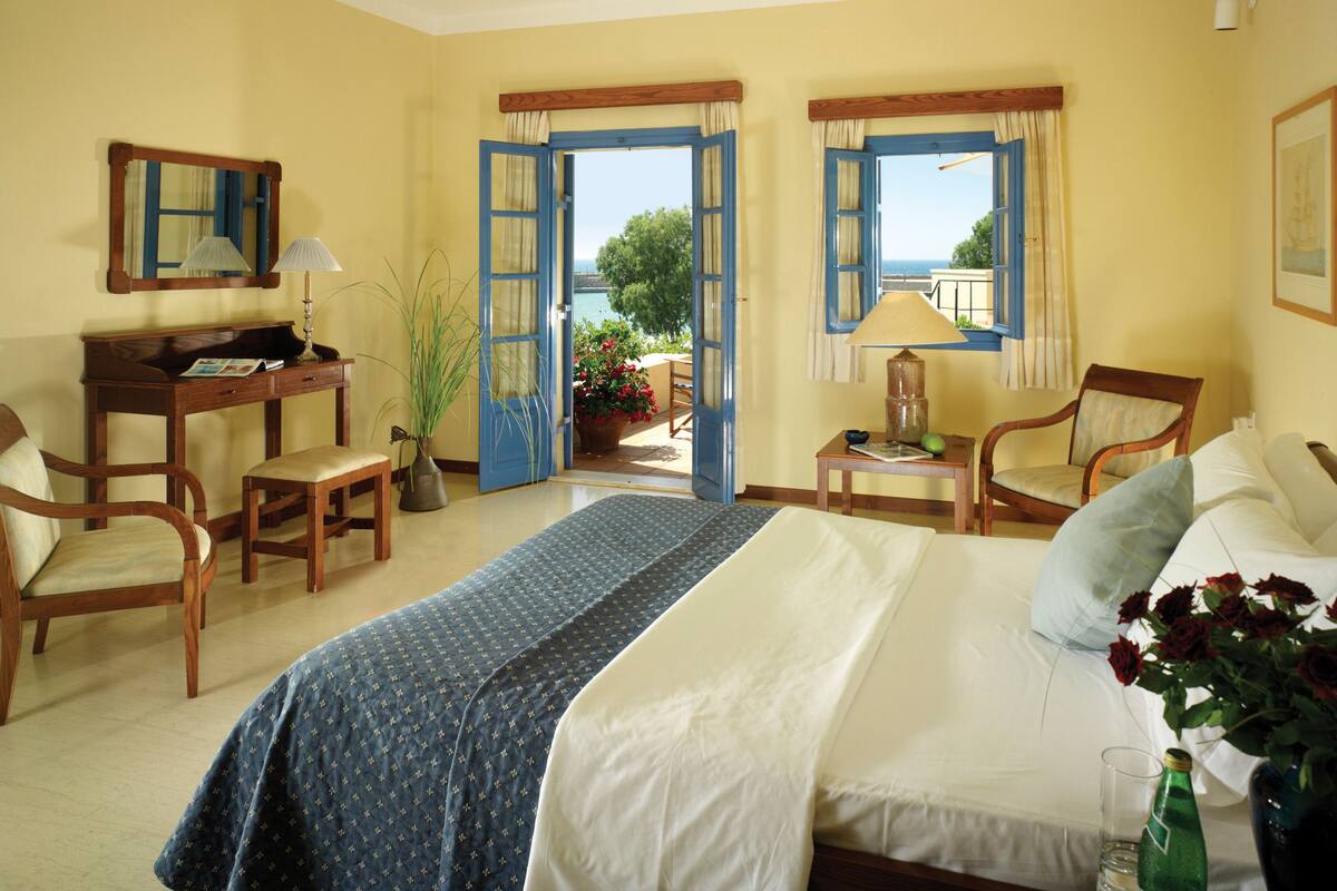 Quarto de hotel com uma cama, duas cadeiras, uma escrivaninha e vista para o exterior.