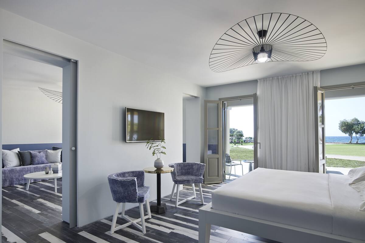 Quarto de hotel moderno com uma cama grande, televisão, área de refeições com cadeiras e vista para o exterior.