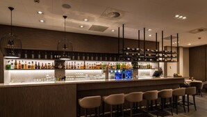 Uma área de bar com um balcão comprido, diversas garrafas, copos e banquetas.