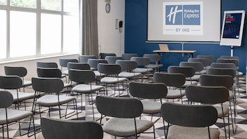 Uma sala de conferências com fileiras de cadeiras cinzas, uma tela de projetor com o logotipo Holiday Inn Express e um relógio na parede.