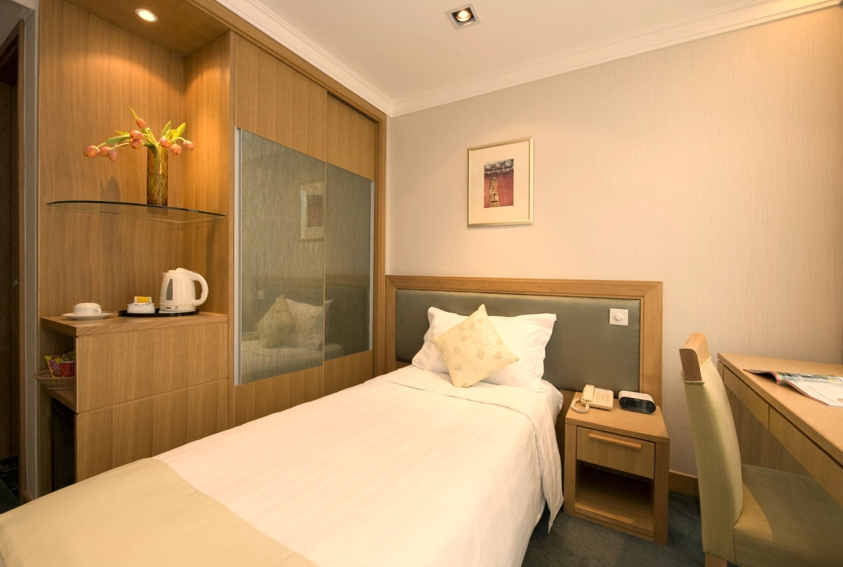 Quarto de hotel com cama, mesa de cabeceira, escrivaninha, cadeira, prateleira de vidro com um vaso e um quadro na parede.
