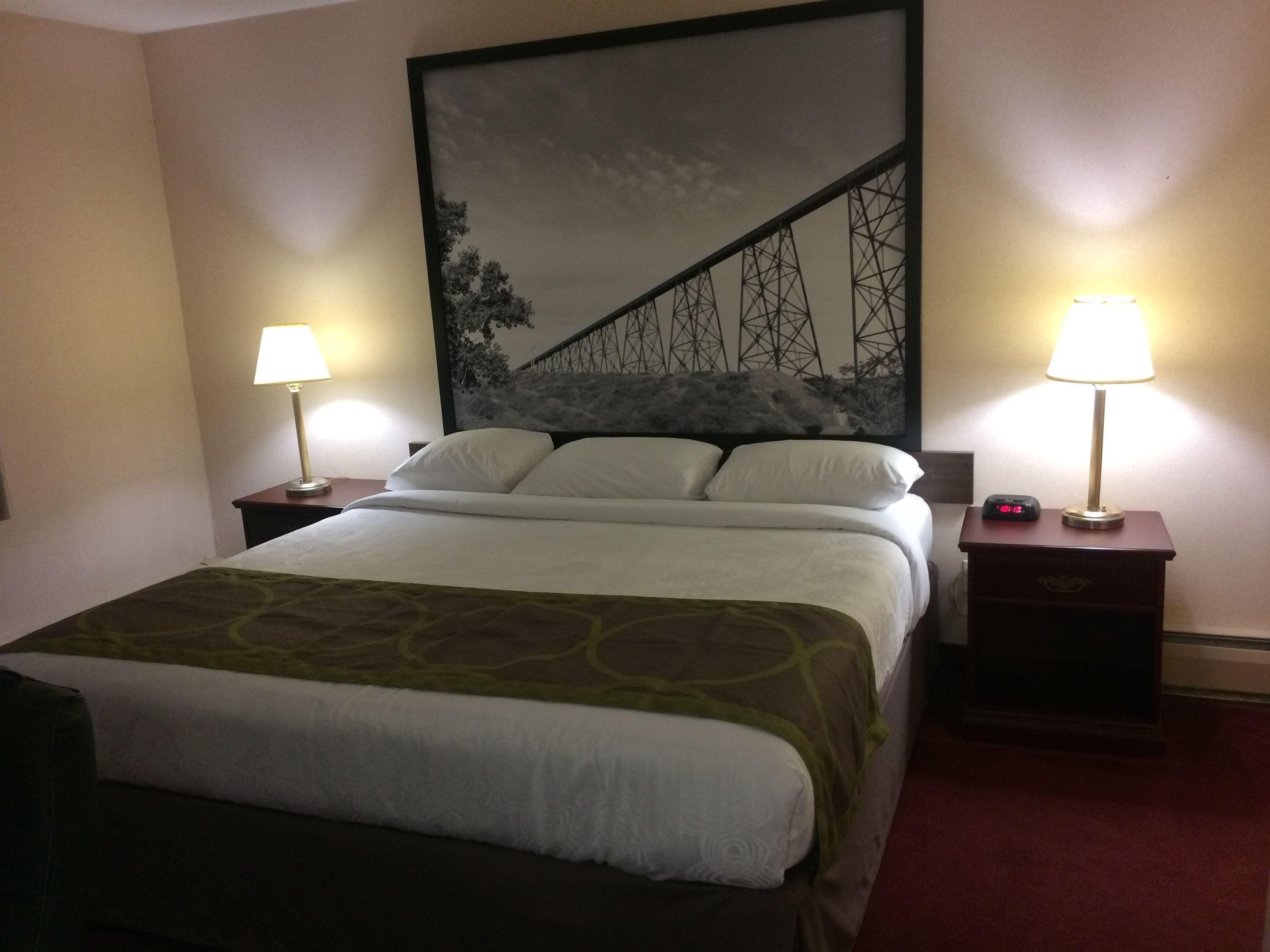 Quarto de hotel com uma cama grande, duas mesas de cabeceira com luminárias, um criado-mudo e um quadro emoldurado na parede.