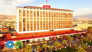 Um hotel grande com telhado vermelho e vários andares, rodeado por palmeiras e um estacionamento.