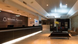 Uma área de recepção de hotel com design moderno, apresentando um balcão de atendimento, espaço de estar e um vaso decorativo com flores.