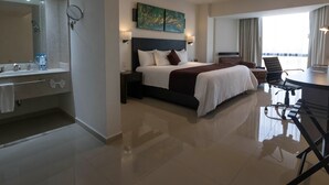 Quarto de hotel com cama, escrivaninha, cadeira, uma janela grande e banheiro com espelho e pia.