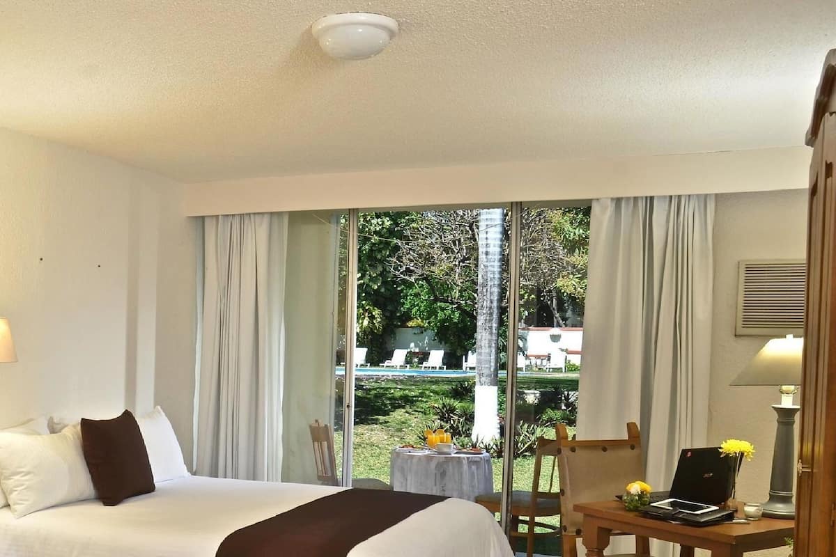 Quarto de hotel com cama, secretária com laptop, cadeira e janela com cortinas.