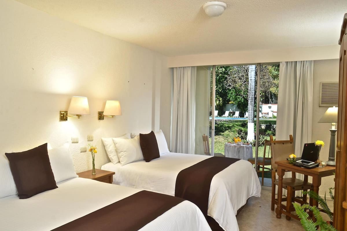 Quarto de hotel com duas camas, uma mesinha com um laptop, uma cadeira e vista para o exterior.
