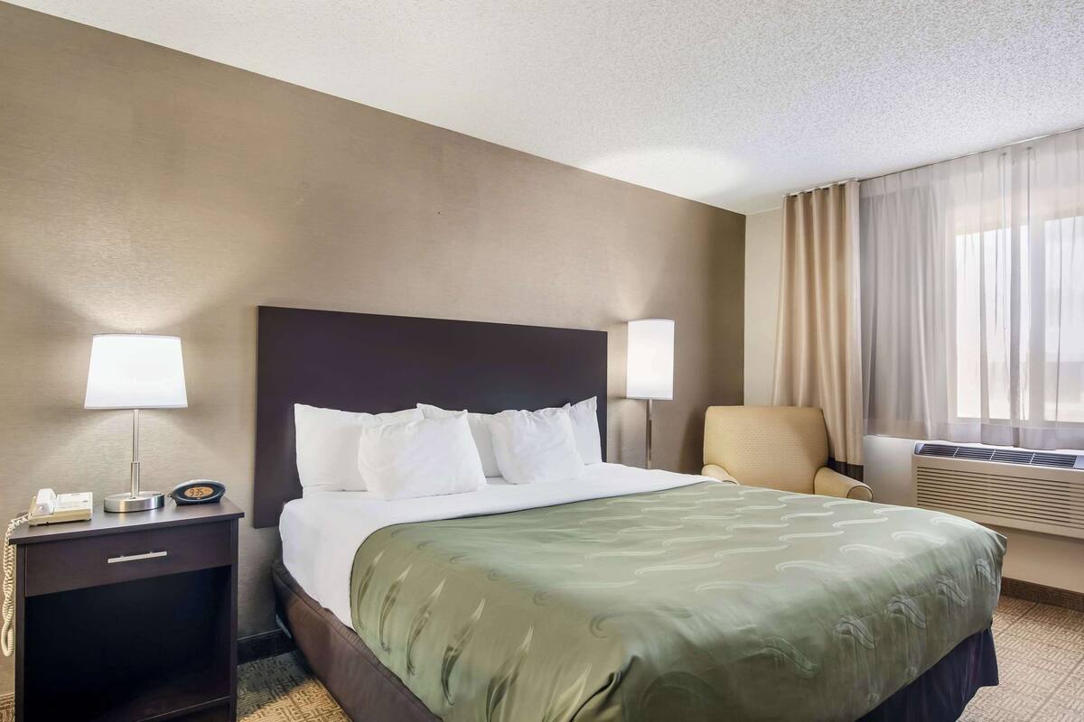 Quarto de hotel com cama, duas luminárias de cabeceira, uma cadeira, ar condicionado e mesa de cabeceira com telefone e relógio.