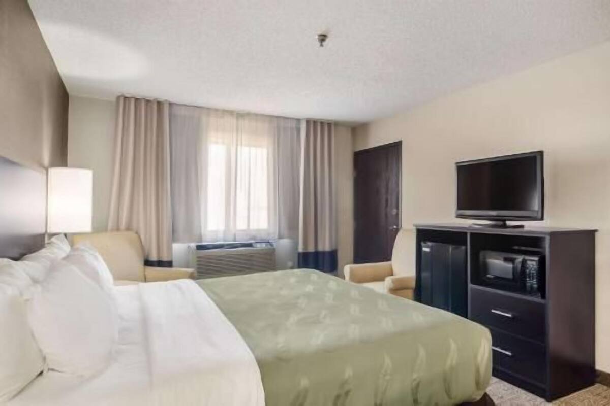 Quarto de hotel com uma cama, televisão em suporte, escrivaninha com cadeira e janela com cortinas.
