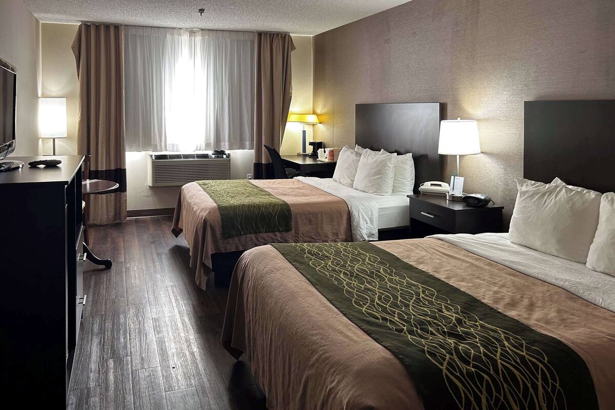 Quarto de hotel com duas camas, uma escrivaninha, uma cadeira, uma luminária e uma janela com cortinas.