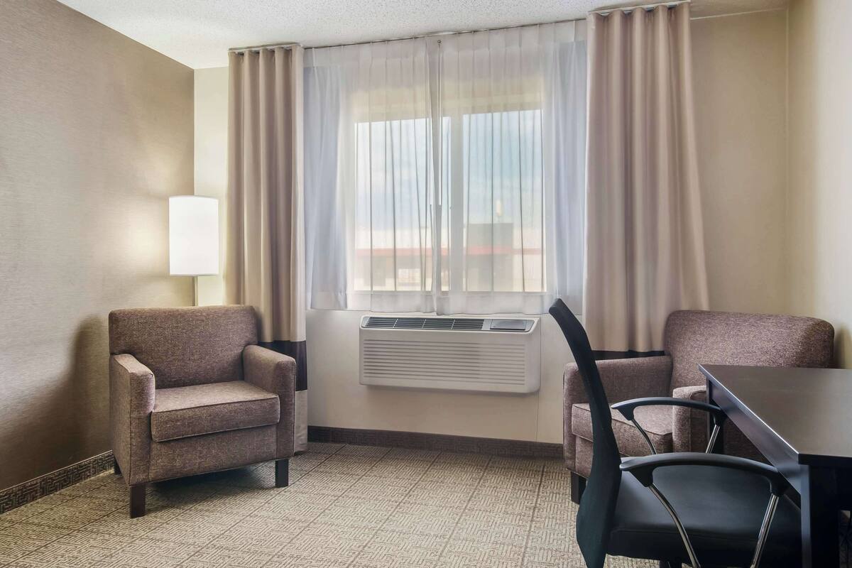 Quarto de hotel com sofá, mesa de jantar e uma cadeira. Há uma janela com cortinas e um aparelho de ar condicionado na parede.