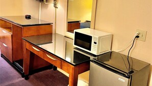 Quarto de hotel pequeno com escrivaninha de madeira, micro-ondas, frigorífico, televisão de ecrã plano e candeeiro de mesa.