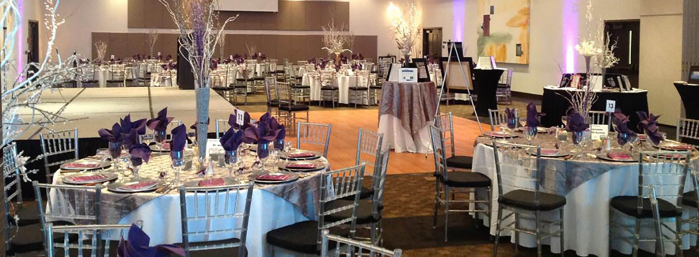 Uma sala de banquetes com mesas redondas preparadas para refeições, cada uma com toalha branca e guardanapos roxos.