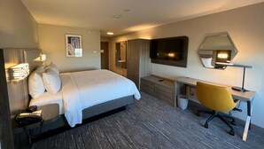 Quarto de hotel com cama, escrivaninha, cadeira, televisão e espelho.