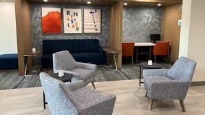 Uma área de lounge moderna com um sofá azul escuro, duas poltronas cinzas e uma pequena mesa. Há obras de arte abstratas nas paredes e uma placa.