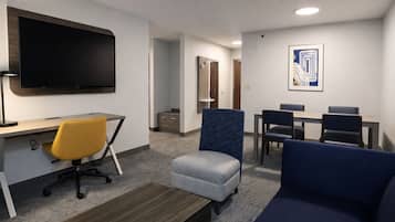 Quarto de hotel com televisão de tela plana na parede, escrivaninha com cadeira, sofá e um pequeno banquinho. Há uma área de refeições com mesa e cadeiras, além de uma pintura abstrata emoldurada na parede.