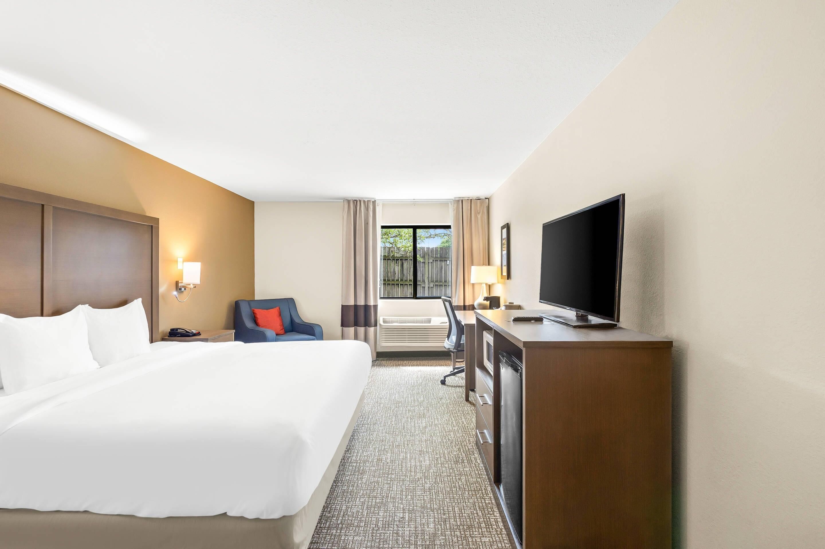 Quarto de hotel com uma cama grande, uma escrivaninha com televisão, uma cadeira e uma janela com cortinas.