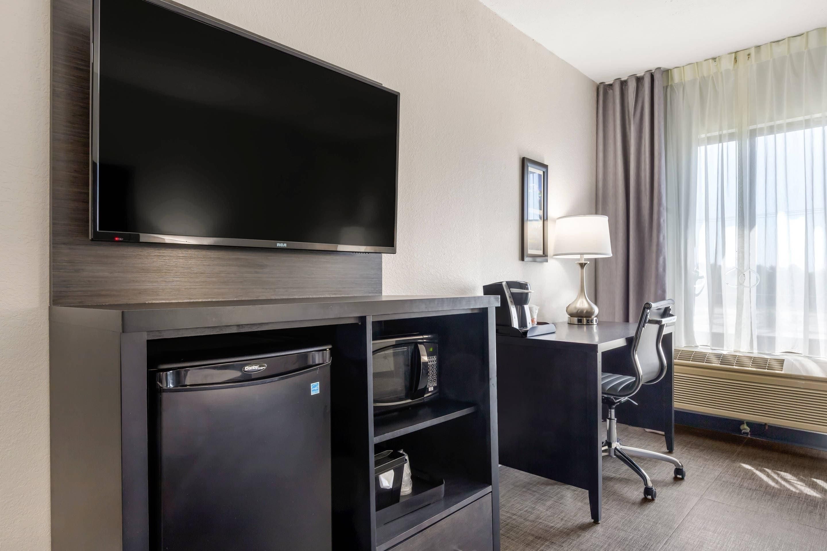 Quarto de hotel com televisão de tela plana, minibar, micro-ondas, escrivaninha com cadeira, luminária e janela com cortinas.