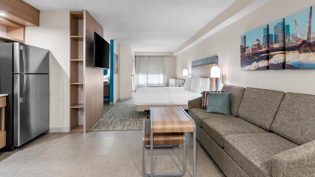 Quarto de hotel com cozinha compacta, cama, sofá, televisão e pintura com vista para a cidade.