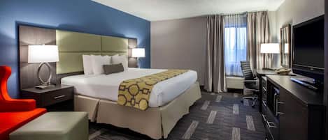 Quarto de hotel com uma cama grande, uma escrivaninha com televisão, uma cadeira vermelha e um pequeno banquinho.