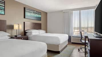 Quarto de hotel com duas camas, uma escrivaninha, uma cadeira e uma janela grande com cortinas.