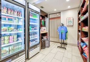 Quarto de hotel com minibar, geladeira abastecida com bebidas, mala e camiseta em um suporte.