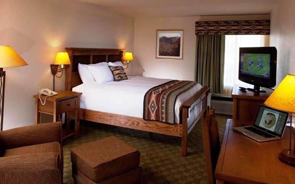 Quarto de hotel com cama, abajures, televisão, laptop, telefone e uma mesinha.