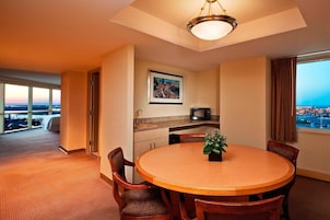 Quarto de hotel com mesa de jantar, cadeiras, pequena cozinha, quadro na parede e vista da cidade por grandes janelas.