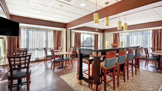 Uma sala de jantar de hotel com mesas e cadeiras de madeira, grandes janelas com cortinas e luminárias pendentes.