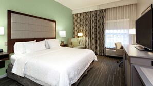 Quarto de hotel com cama grande, escrivaninha, televisão de tela plana, uma cadeira e janela com cortinas.