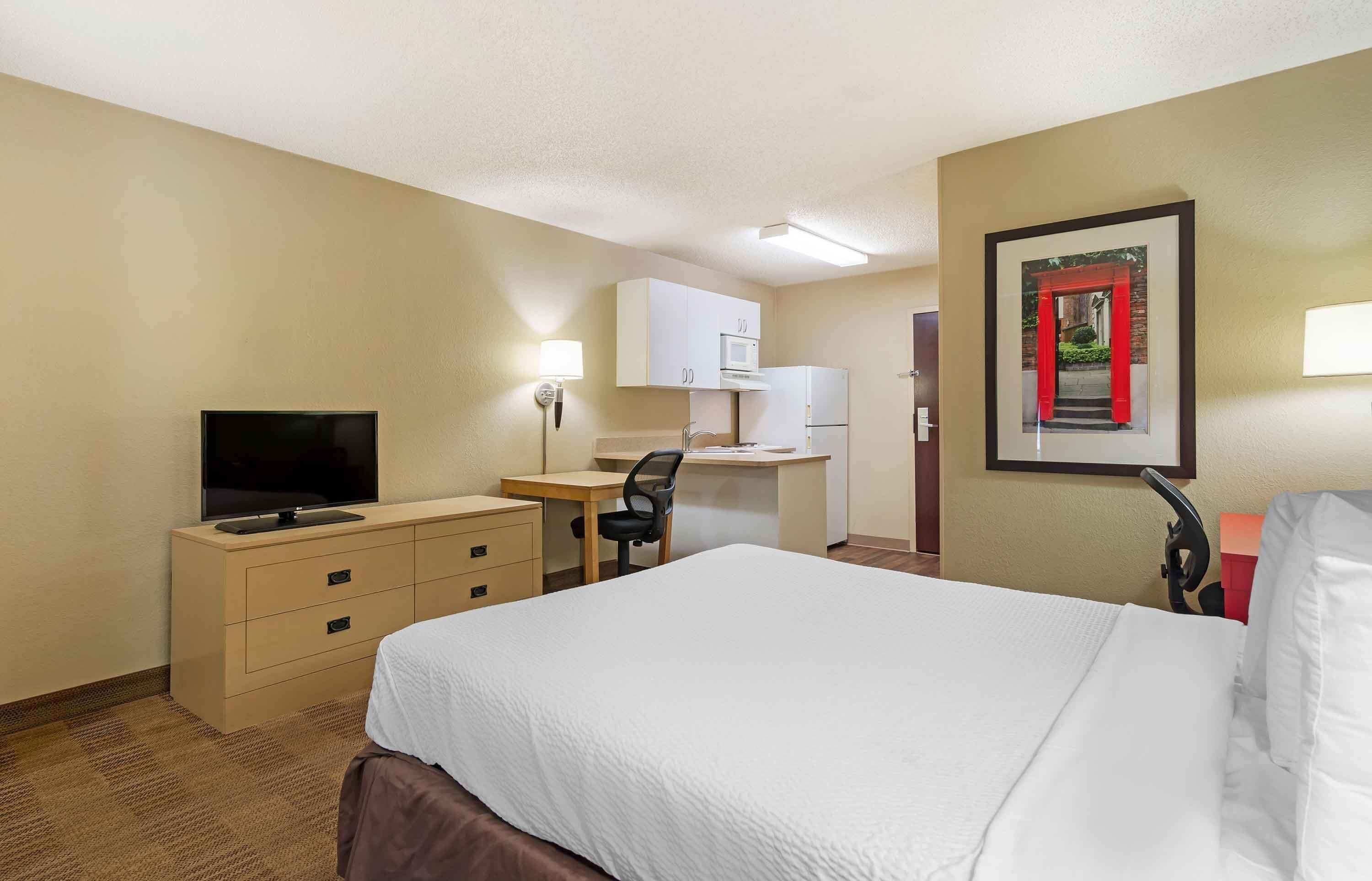 Quarto de hotel com cama, escrivaninha com cadeira, televisão, micro-ondas e frigorífico.