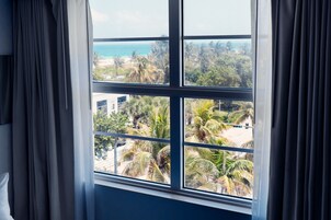 Quarto com uma janela grande que oferece vista para palmeiras e uma praia.
