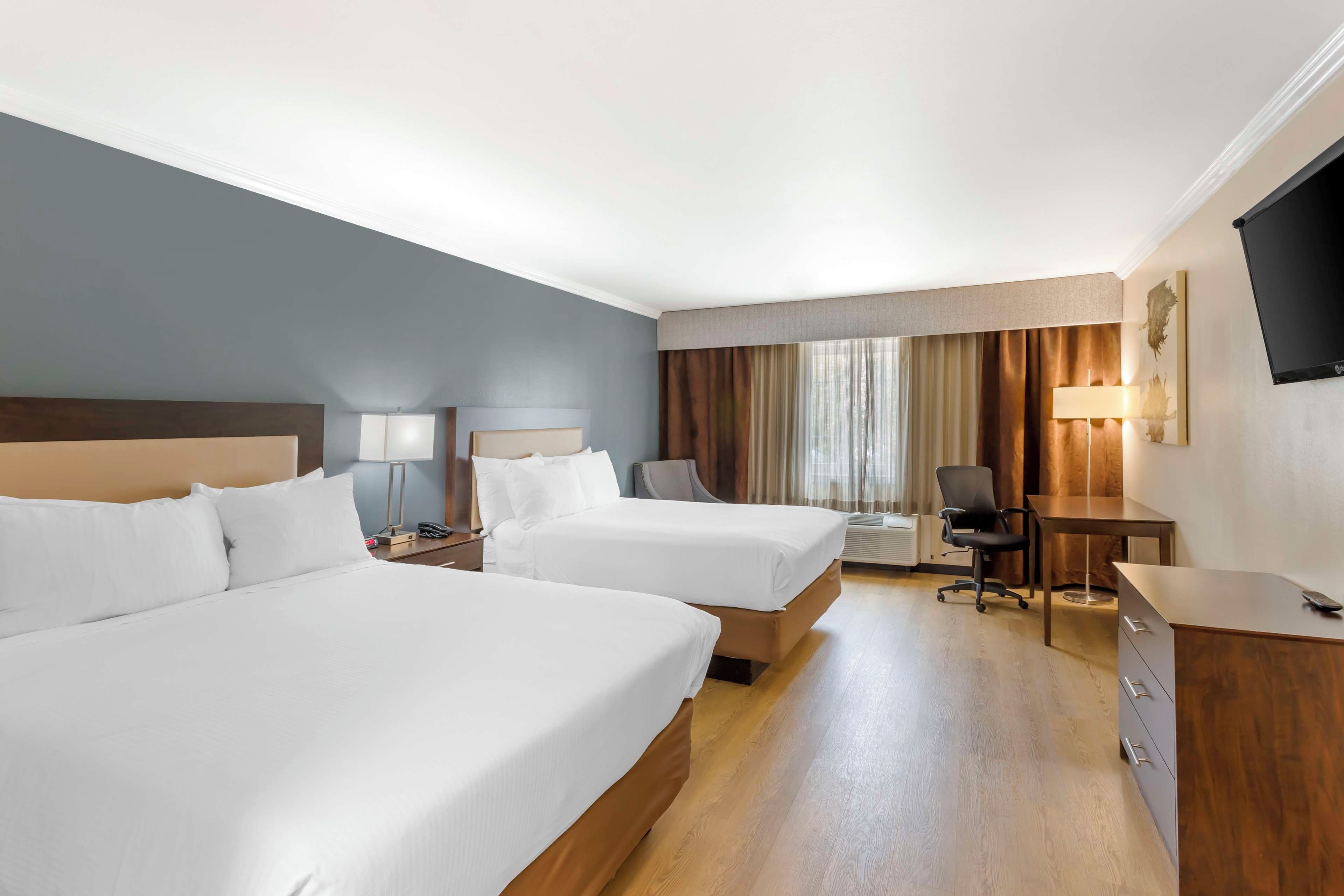 Quarto de hotel com duas camas, uma escrivaninha, uma cadeira, uma televisão e uma janela com cortinas.