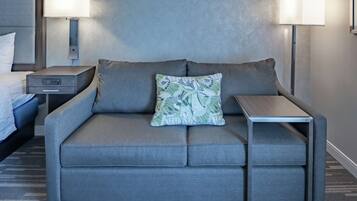 Quarto de hotel moderno com sofá cinza, almofada estampada, mesa de cabeceira e abajur.