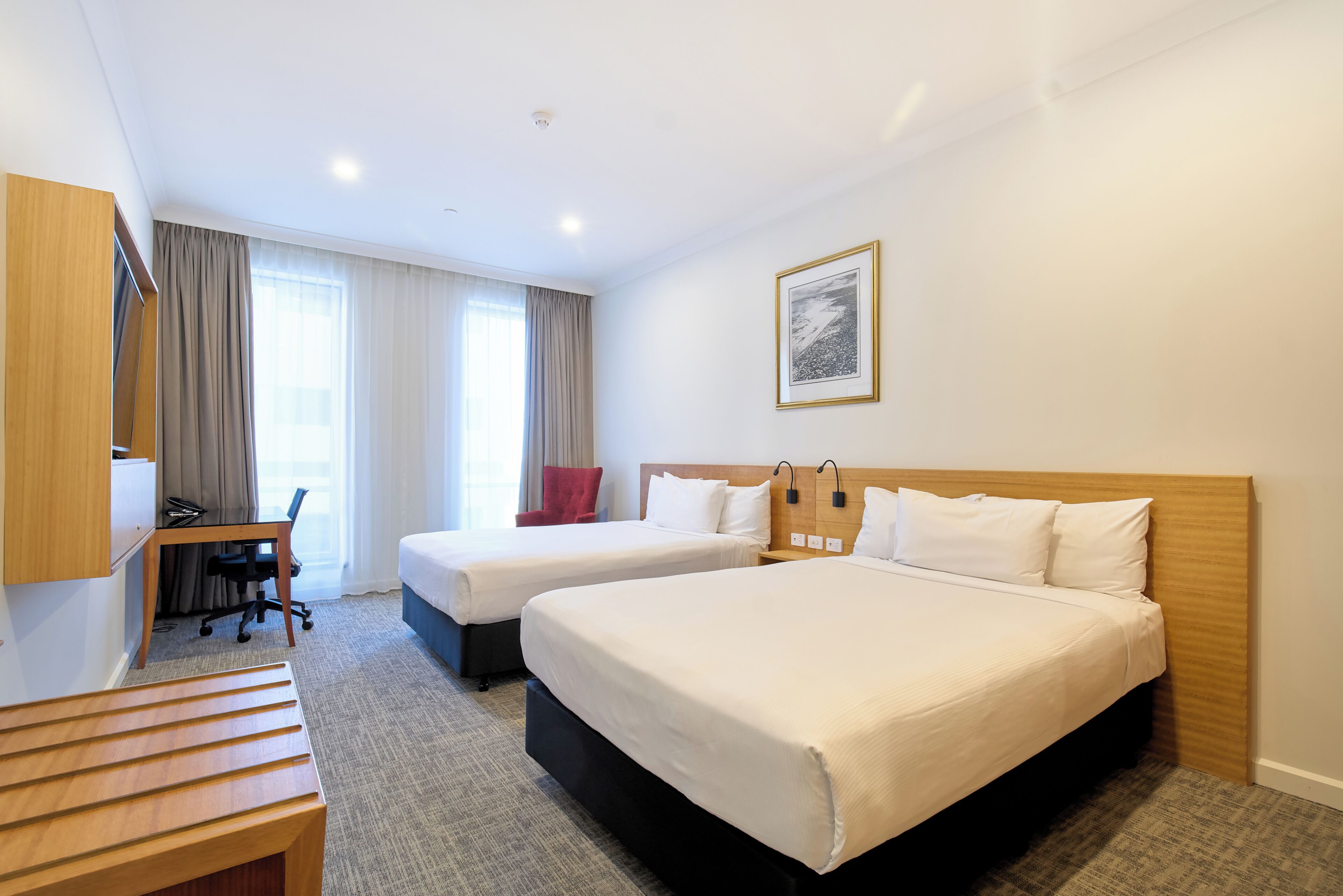 Quarto de hotel com duas camas, uma escrivaninha, uma cadeira e uma janela grande com cortinas.