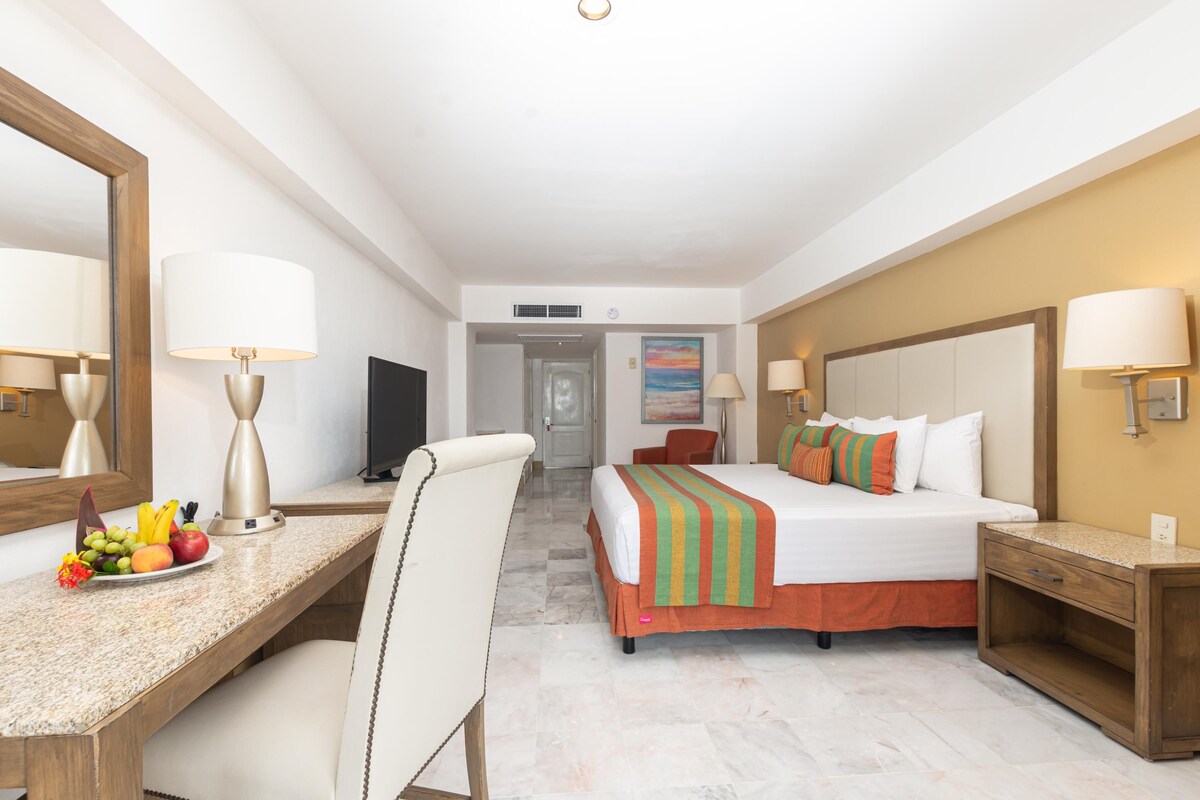 Quarto de hotel com cama, escrivaninha, cadeira e um espelho grande.
