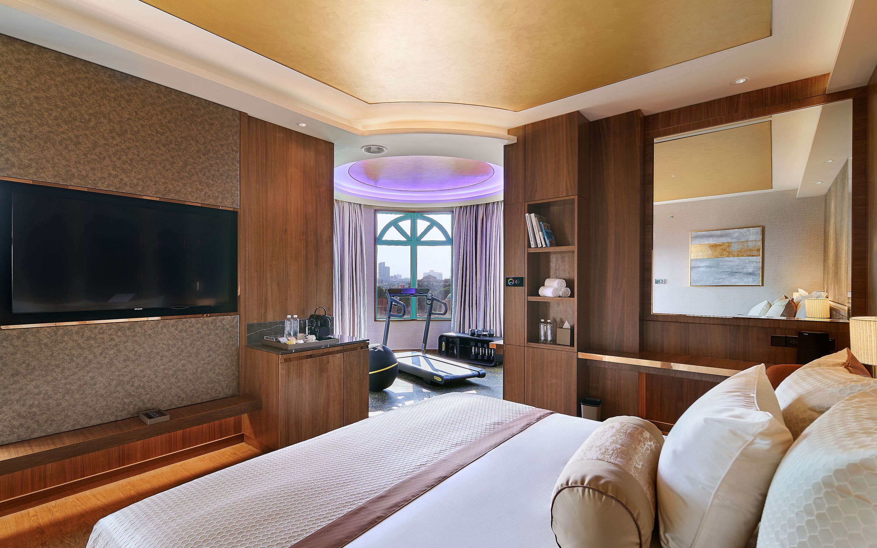 Quarto de hotel com cama grande, televisão, esteira e vista para a cidade pela janela.