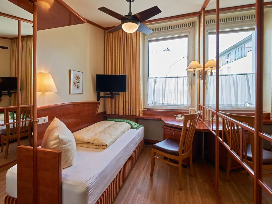 Quarto de hotel pequeno com uma cama de solteiro, uma mesa, uma cadeira, uma televisão e uma janela com cortinas.