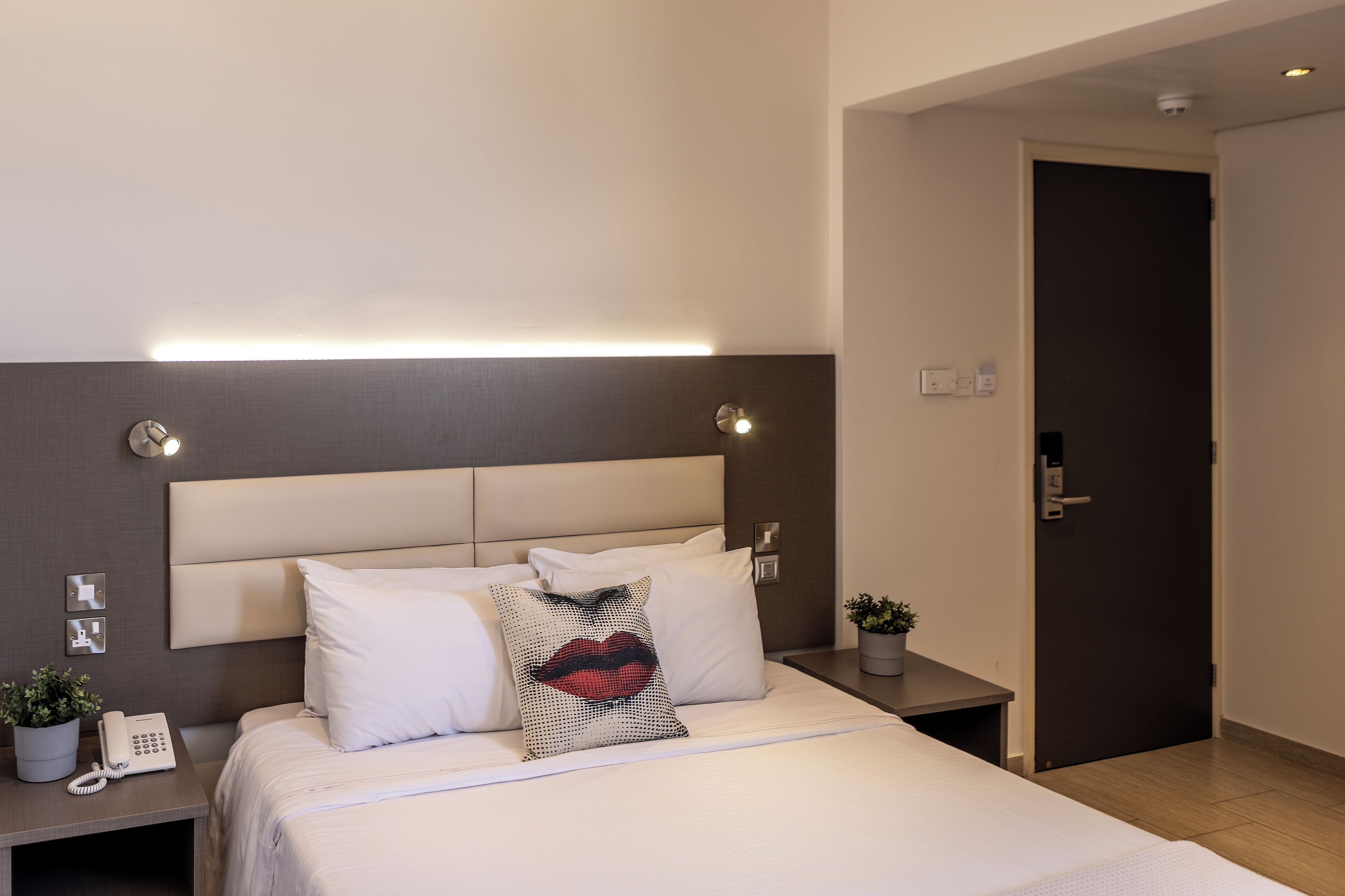 Quarto de hotel moderno com cama bem arrumada, telefone ao lado da cama, uma pequena planta em vaso e porta à direita.