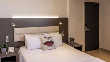 Quarto de hotel moderno com cama bem arrumada, telefone ao lado da cama, uma pequena planta em vaso e porta à direita.