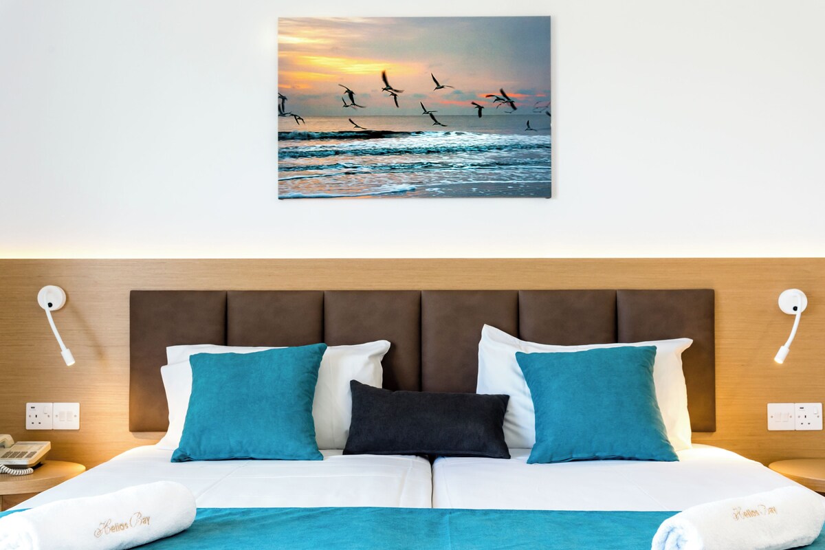Quarto de hotel com uma cama grande, duas almofadas, uma mesinha de cabeceira, um telefone e um quadro na parede com pássaros voando sobre o oceano.