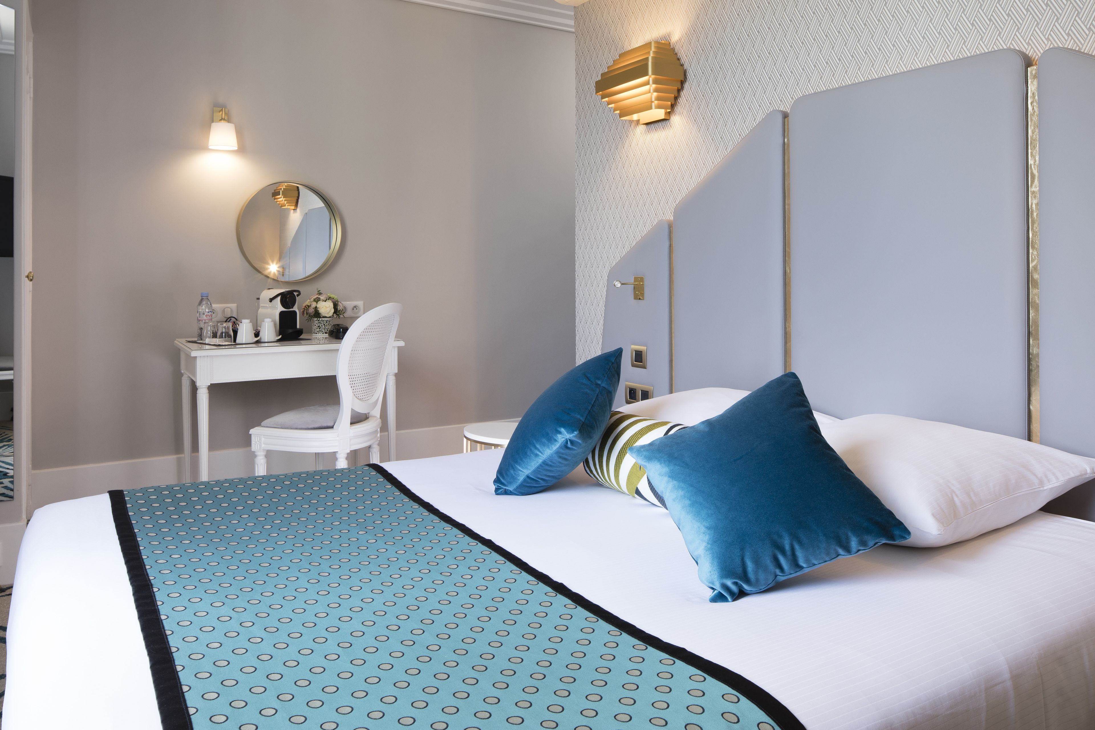 Quarto de hotel com uma cama grande, uma escrivaninha com cadeira, um espelho e uma luminária na parede.