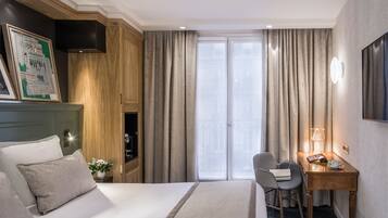 Quarto de hotel com cama, escrivaninha, cadeira, luminária e uma grande janela com cortinas.