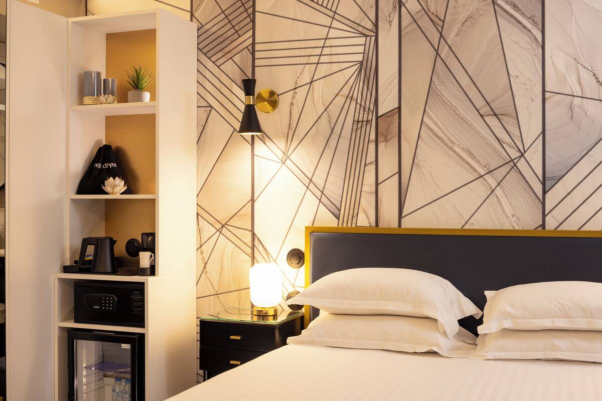 Quarto de hotel com cama, mesa de cabeceira, prateleira com abajur, pequeno frigorífico e parede decorativa com padrões geométricos.