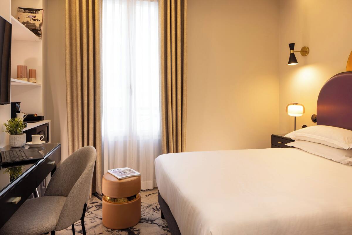 Quarto de hotel com cama, escrivaninha com computador, cadeira, mesa pequena e janela com cortinas.