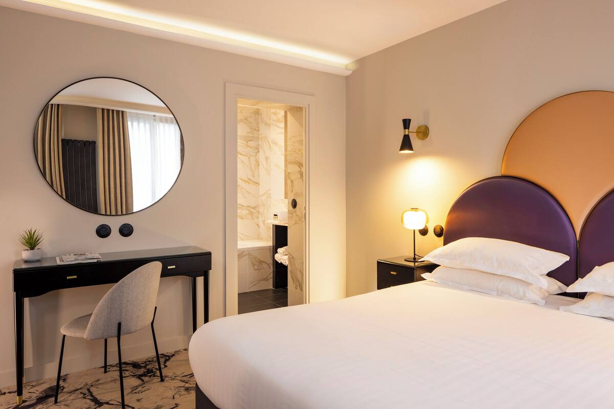 Quarto de hotel com uma cama, uma escrivaninha com cadeira, um espelho redondo, uma mesa de cabeceira com abajur e uma porta de banheiro.