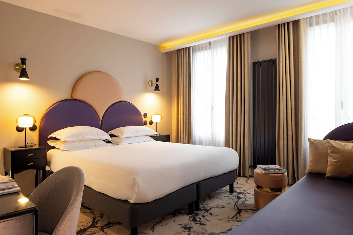 Quarto de hotel com uma cama grande, duas mesas de cabeceira, uma cadeira, uma mesinha e uma janela com cortinas.