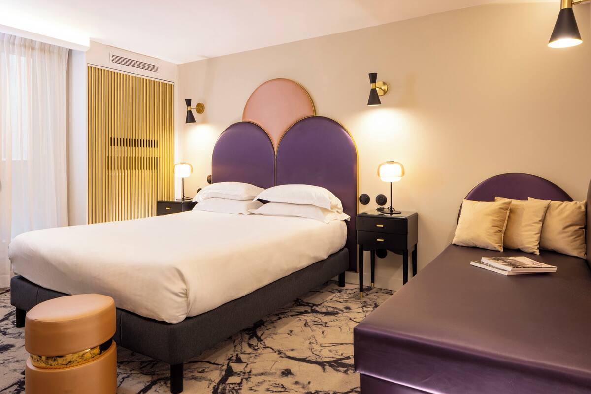 Quarto de hotel com uma cama grande, uma cama menor, uma mesa de cabeceira com abajur e um painel decorativo na parede.