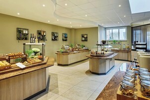 Espaço de café da manhã de hotel, com diversas estações de serviço, buffet com vários tipos de comida e estação de café.