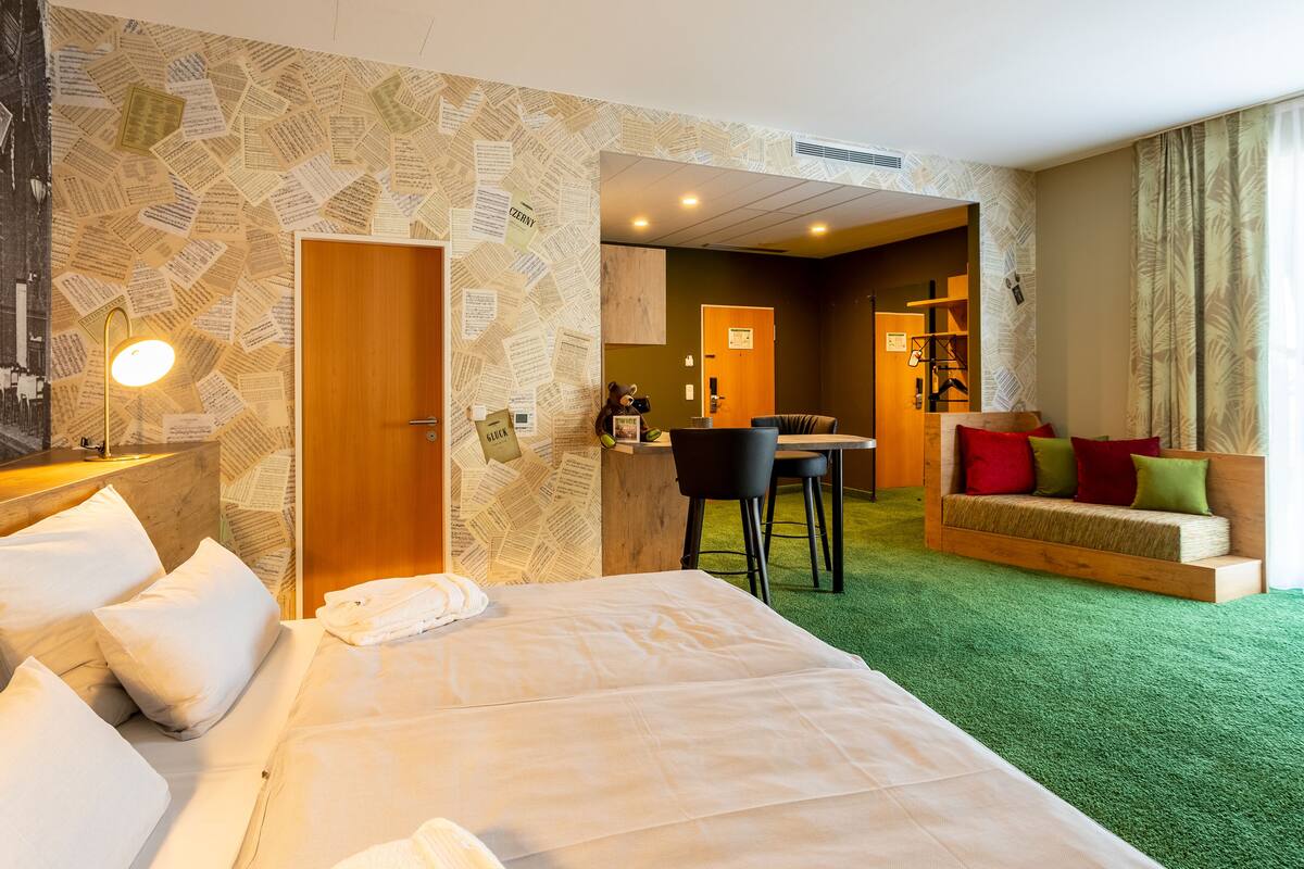 Quarto de hotel com cama, escrivaninha, cadeira, sofá e parede coberta de recortes de jornal.
